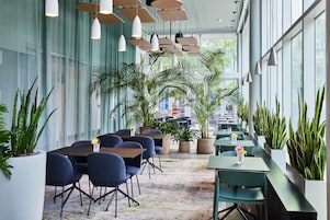 Uma área de refeições interna moderna, com mesas, cadeiras, grandes janelas e luminárias pendentes.