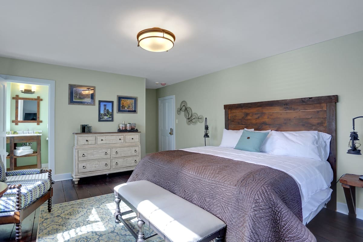 Quarto com cama grande, cabeceira de madeira, um banco, cômoda e espelho.
