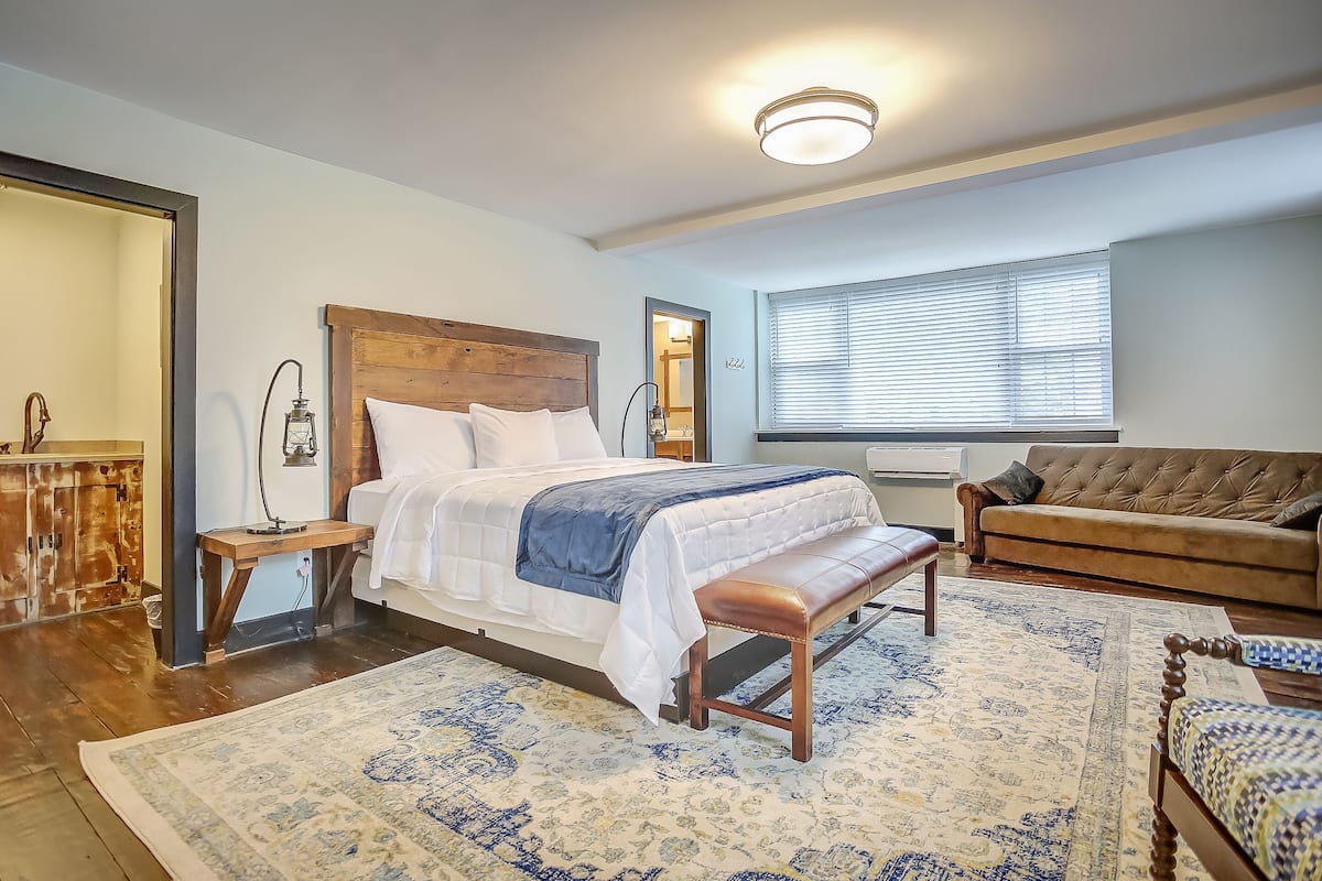 Quarto com uma cama grande, cabeceira de madeira, um banquinho, um sofá e um tapete estampado.