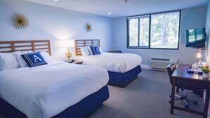 Quarto de hotel com duas camas, uma escrivaninha, uma cadeira, uma televisão e uma janela com persianas.