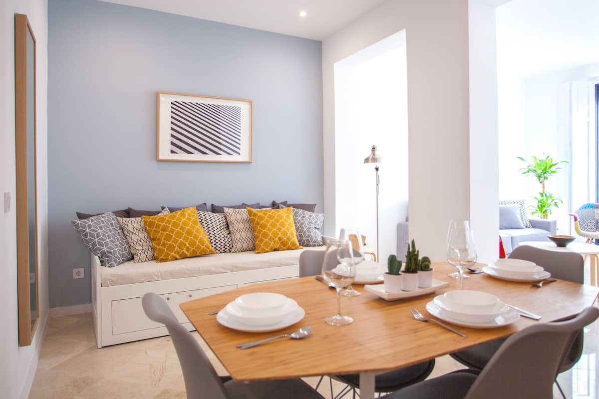 Uma sala de jantar moderna com uma mesa de madeira posta para quatro pessoas, um sofá com almofadas estampadas e uma obra de arte emoldurada na parede.