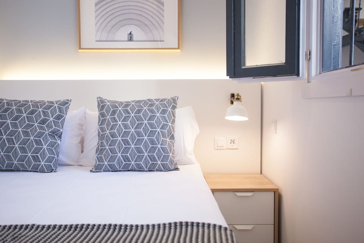 Uma cama bem arrumada com almofadas estampadas, uma mesa de cabeceira com abajur e um quadro na parede.