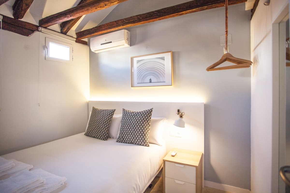 Quarto com uma cama, duas almofadas estampadas, mesa de cabeceira com abajur, cabide de roupa e um quadro na parede.