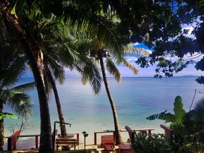 Vista da praia com palmeiras, um banco de madeira e um corrimão com vista para o mar.