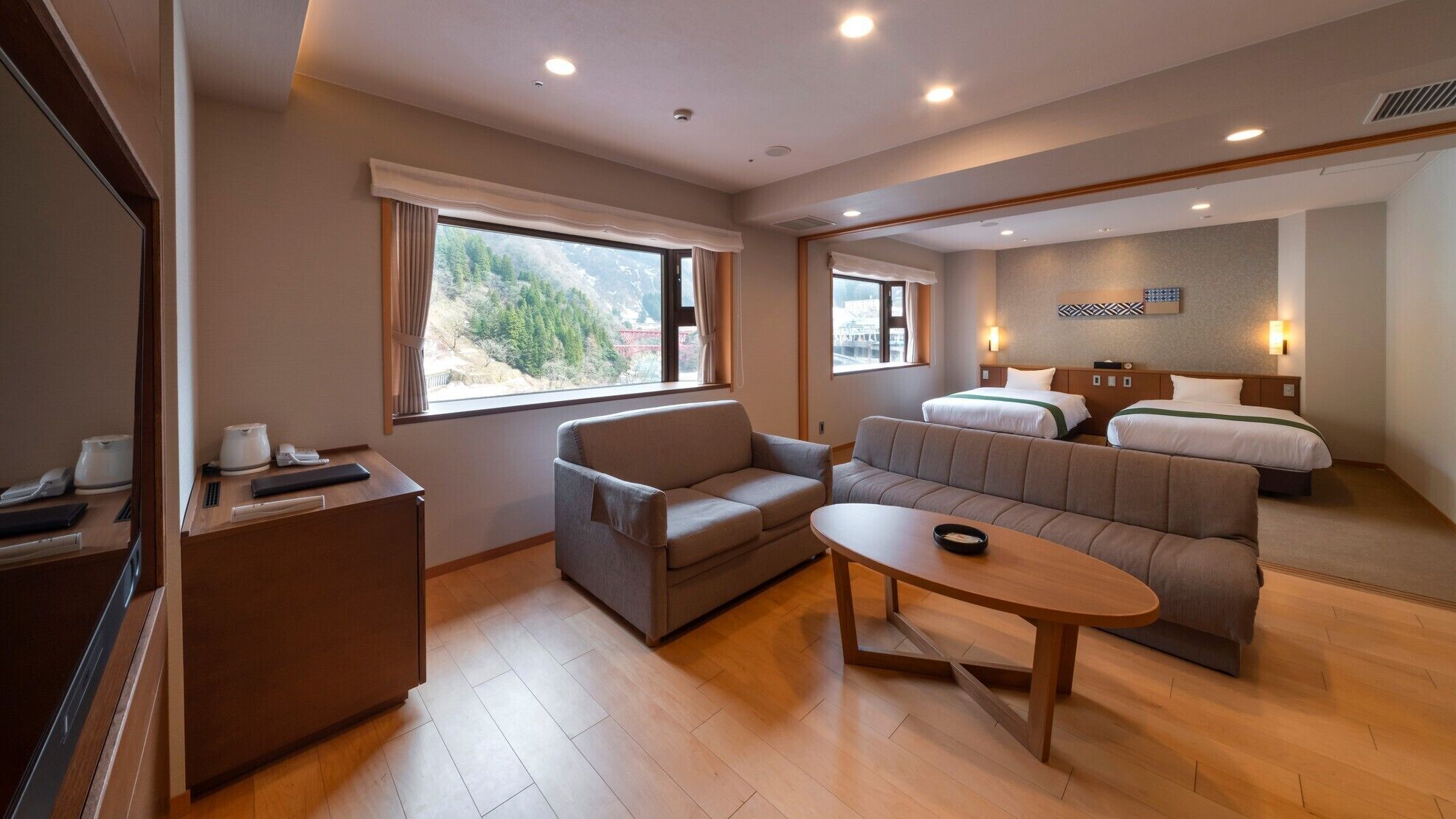 Quarto de hotel com sofá, uma pequena mesa, escrivaninha com telefone e abajur, cama com duas almofadas e vista para uma floresta pela janela.