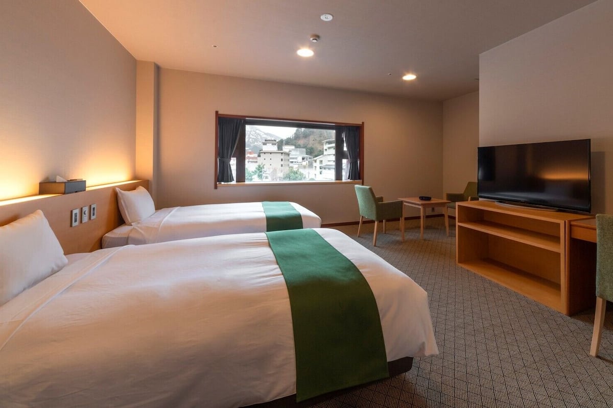Quarto de hotel com duas camas, televisão, escrivaninha e janela com vista para edifícios.