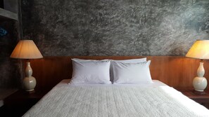Cama bem arrumada com lençóis e almofadas brancas, com duas luminárias laterais sobre mesas de cabeceira de madeira.