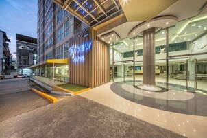 Entrada de hotel com design moderno, destacando fachada de vidro, uma coluna central e interior bem iluminado.