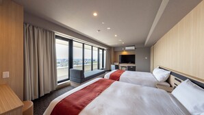 Quarto de hotel com duas camas, uma escrivaninha, televisão e uma janela grande com vista para a cidade.