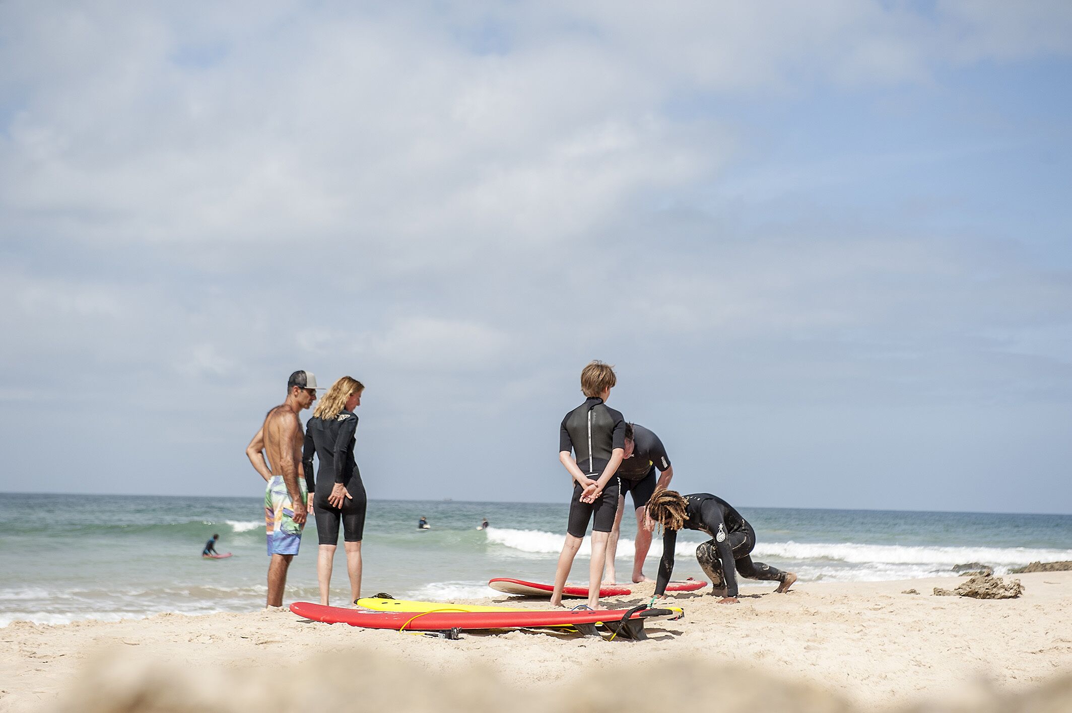 Uma praia com pessoas em pé e sentadas na areia, algumas vestindo fatos de neoprene, e pranchas de surf alinhadas na praia.
