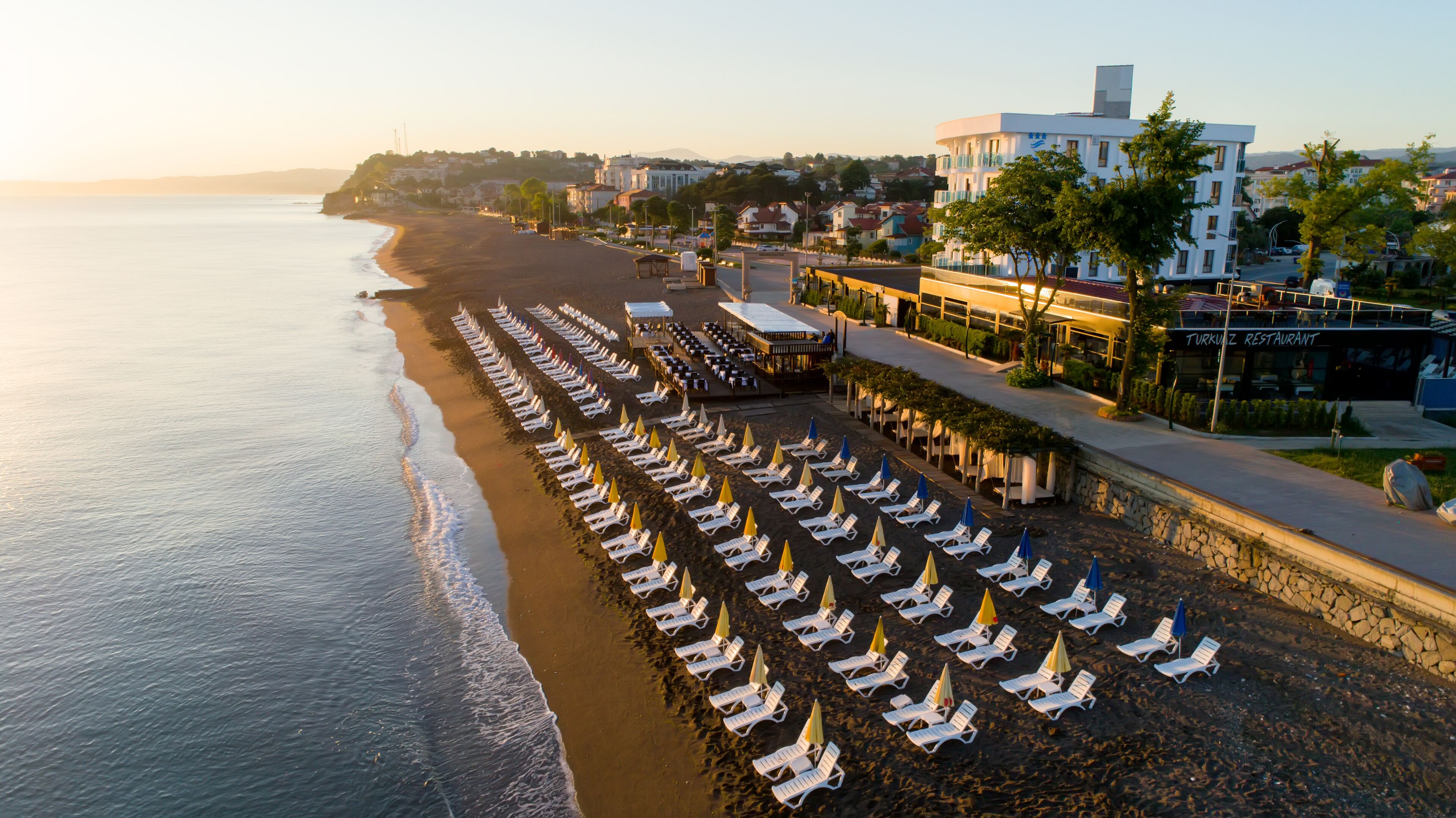 Um hotel à beira-mar com fileiras de espreguiçadeiras e guarda-sóis, um restaurante e vista para o mar.