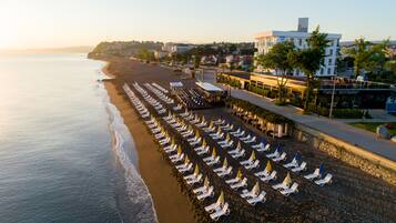 Um hotel à beira-mar com fileiras de espreguiçadeiras e guarda-sóis, um restaurante e vista para o mar.