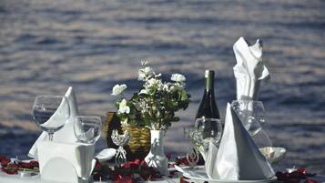Mesa posta para dois com taças de vinho, uma garrafa e um vaso de flores, com vista para um corpo de água.