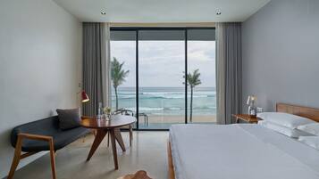 Quarto de hotel com cama, escrivaninha, cadeira, mesa pequena com abajur e vista para o mar.