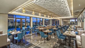 Um restaurante amplo com decoração moderna, apresentando cadeiras de veludo azul, piso de mármore e teto com padrões geométricos.