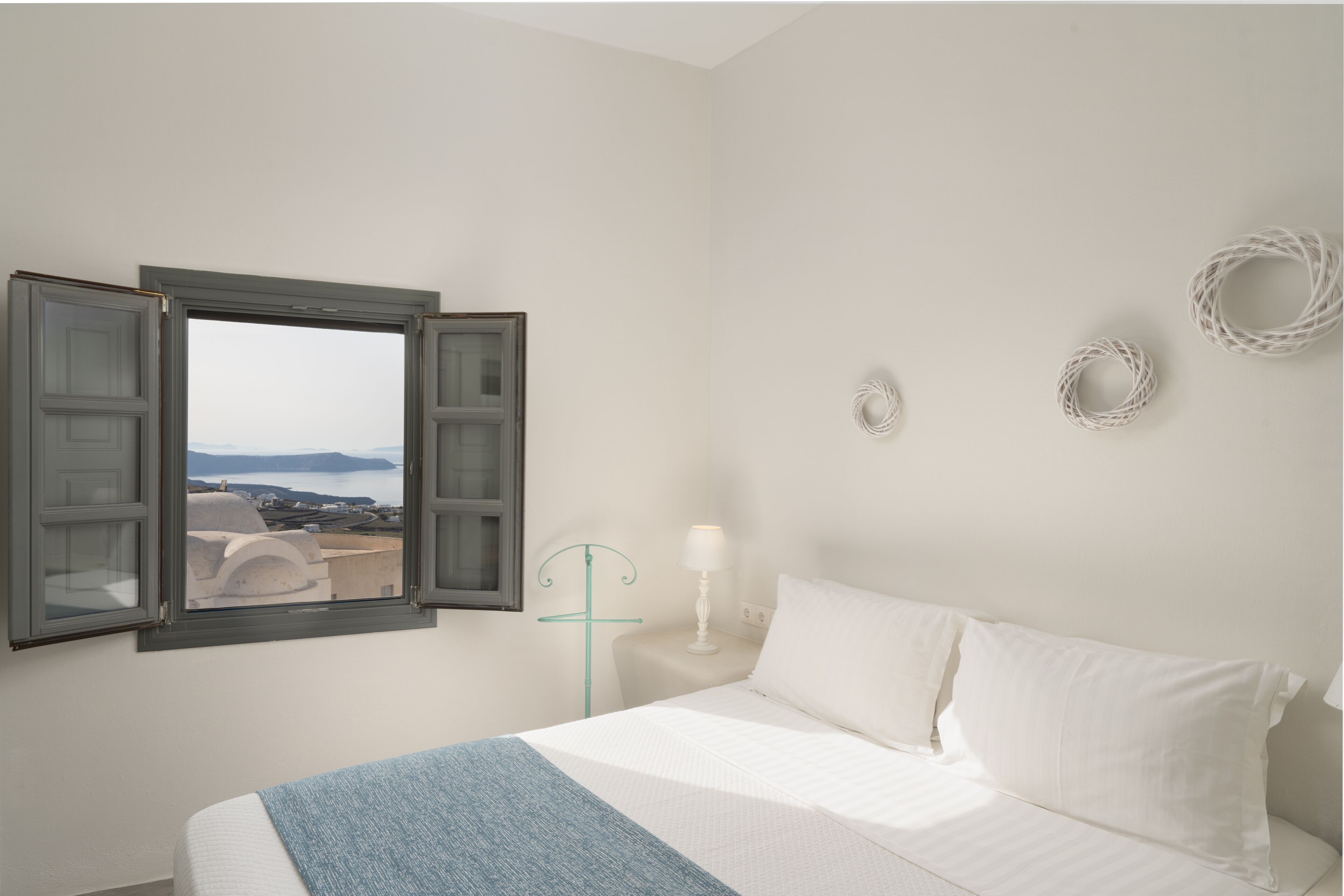Quarto com cama, janela com persiana aberta, mesa de cabeceira com abajur e vista para o mar e edifícios.