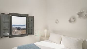 Quarto com cama, janela com persiana aberta, mesa de cabeceira com abajur e vista para o mar e edifícios.