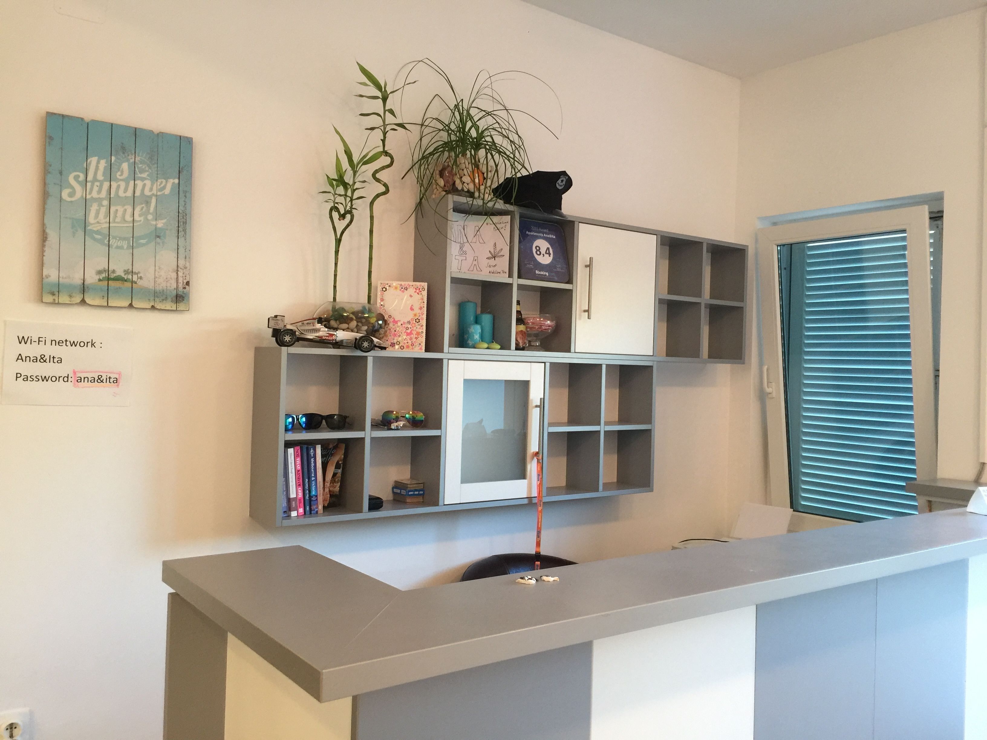 Uma receção com um sinal a indicar a rede Wi-Fi e a password. Há uma estante com objetos decorativos, uma pequena planta e um quadro emoldurado.