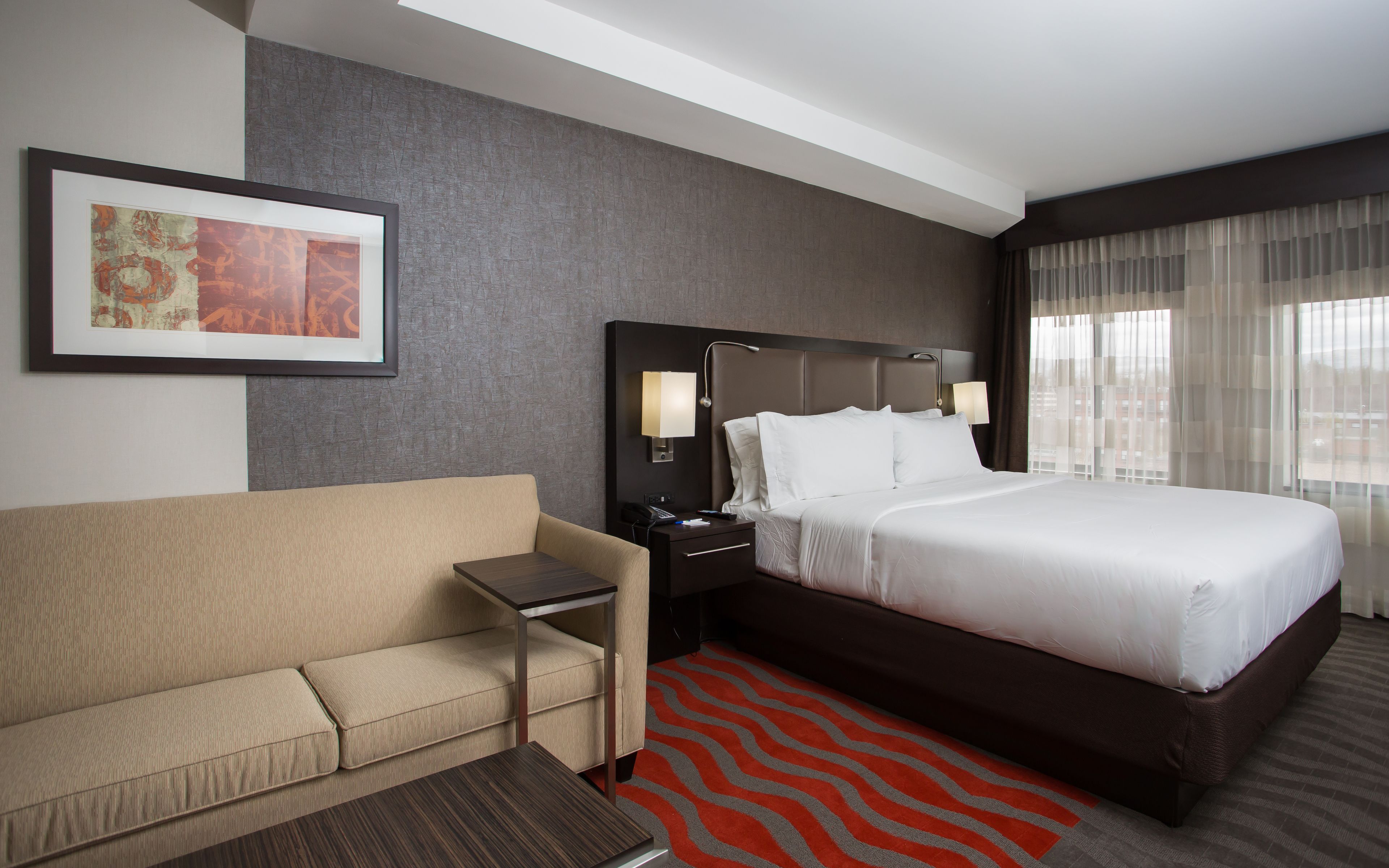 Quarto de hotel com cama, sofá, mesa de cabeceira, luminária e um quadro na parede.