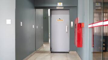 Um corredor com uma porta cinza, um quadro de alarme de incêndio vermelho e uma placa indicando um banheiro.