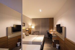 Quarto de hotel com uma cama, uma escrivaninha, uma cadeira, uma televisão e um abajur.