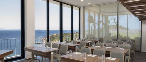 Sala de conferências com vista para o mar, equipada com mesas, cadeiras e garrafas de água.