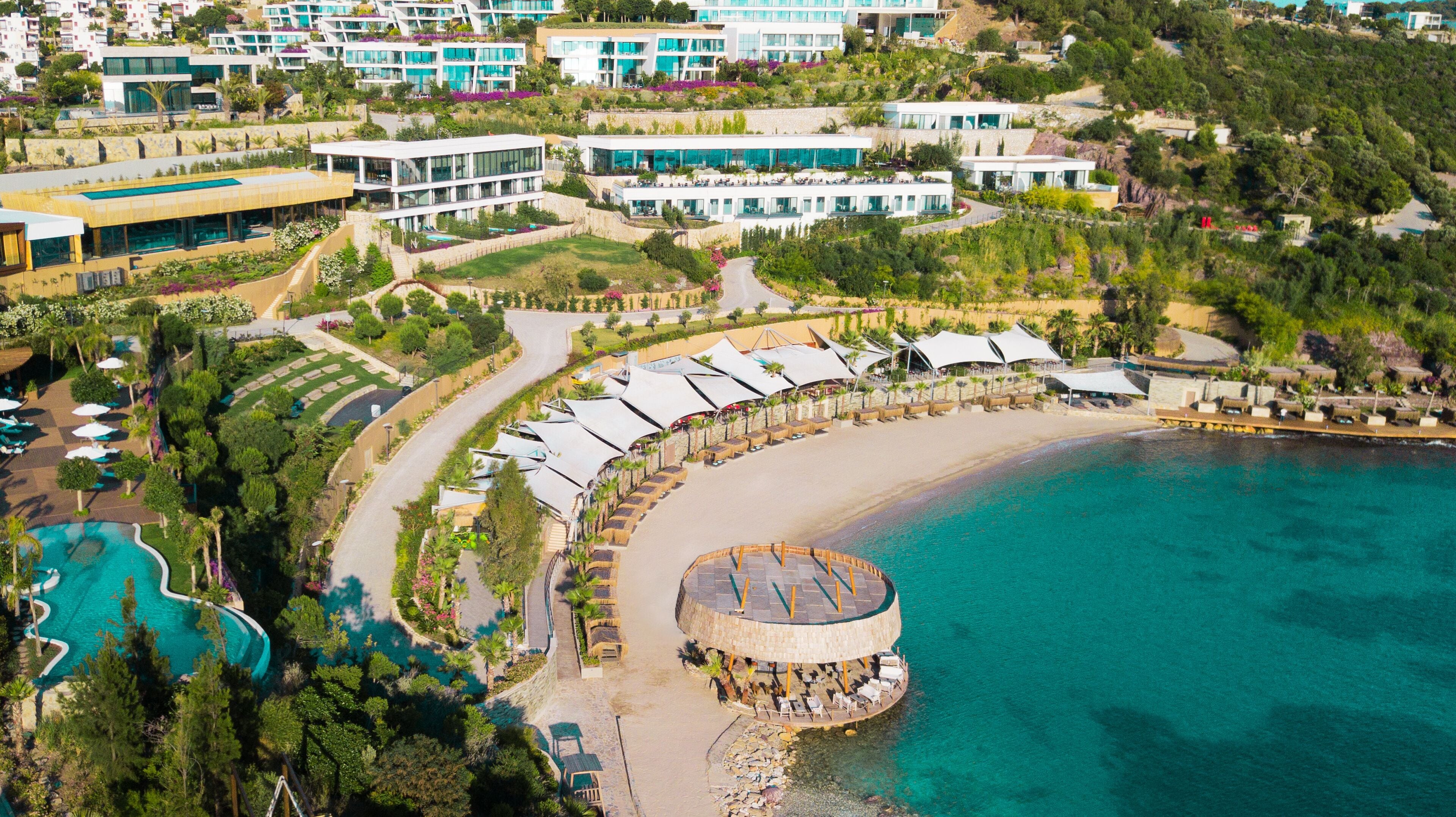 Resort com piscina circular central, área de praia com guarda-sóis e edifícios modernos ao fundo.