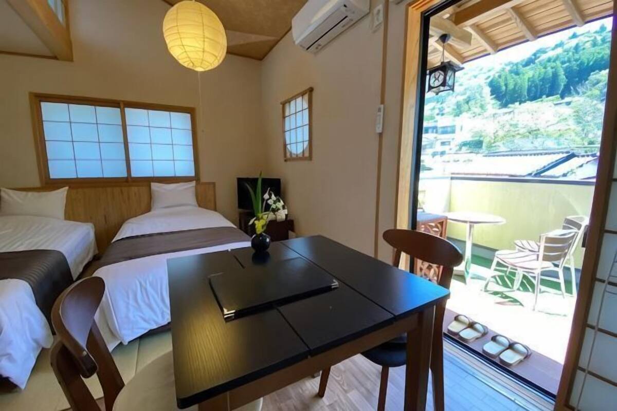 Quarto com cama, mesa com cadeiras, uma mesinha com um vaso e vista para uma varanda com mobiliário exterior.