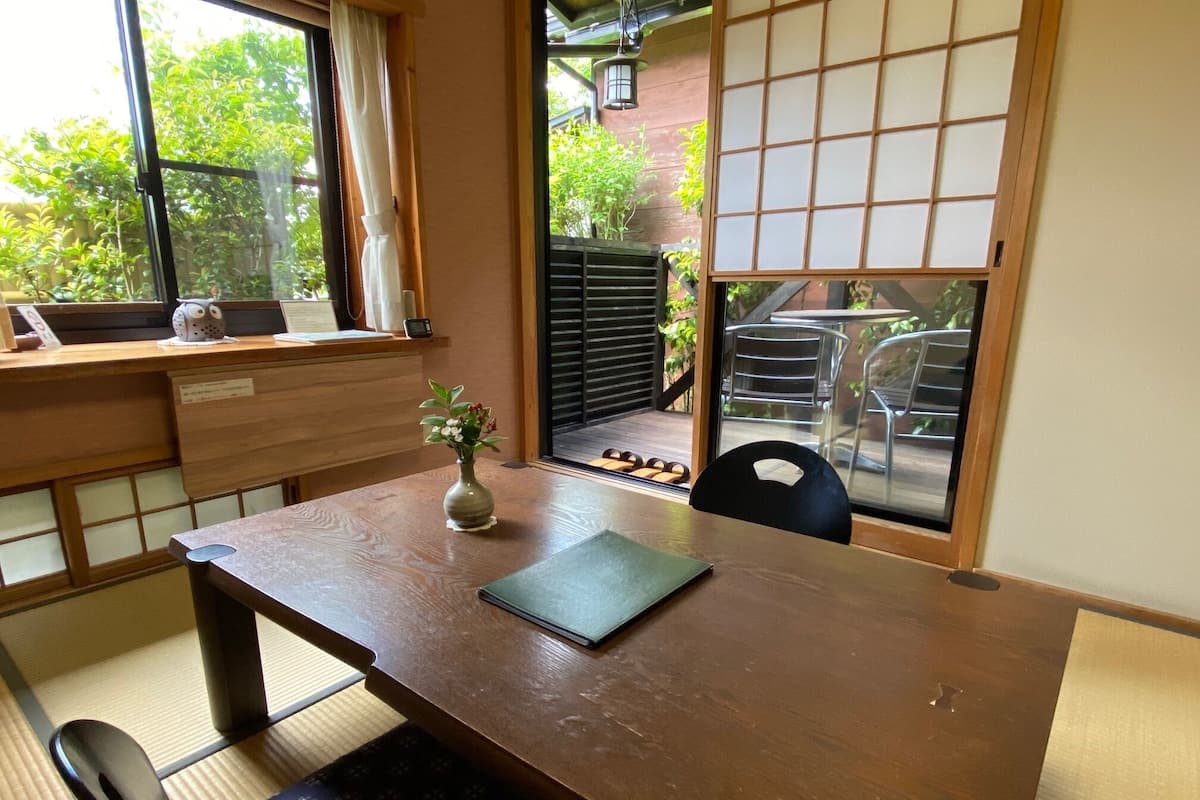 Quarto tradicional em estilo japonês com piso de tatame, uma mesa de madeira com um vaso de flores e vista para a varanda externa.