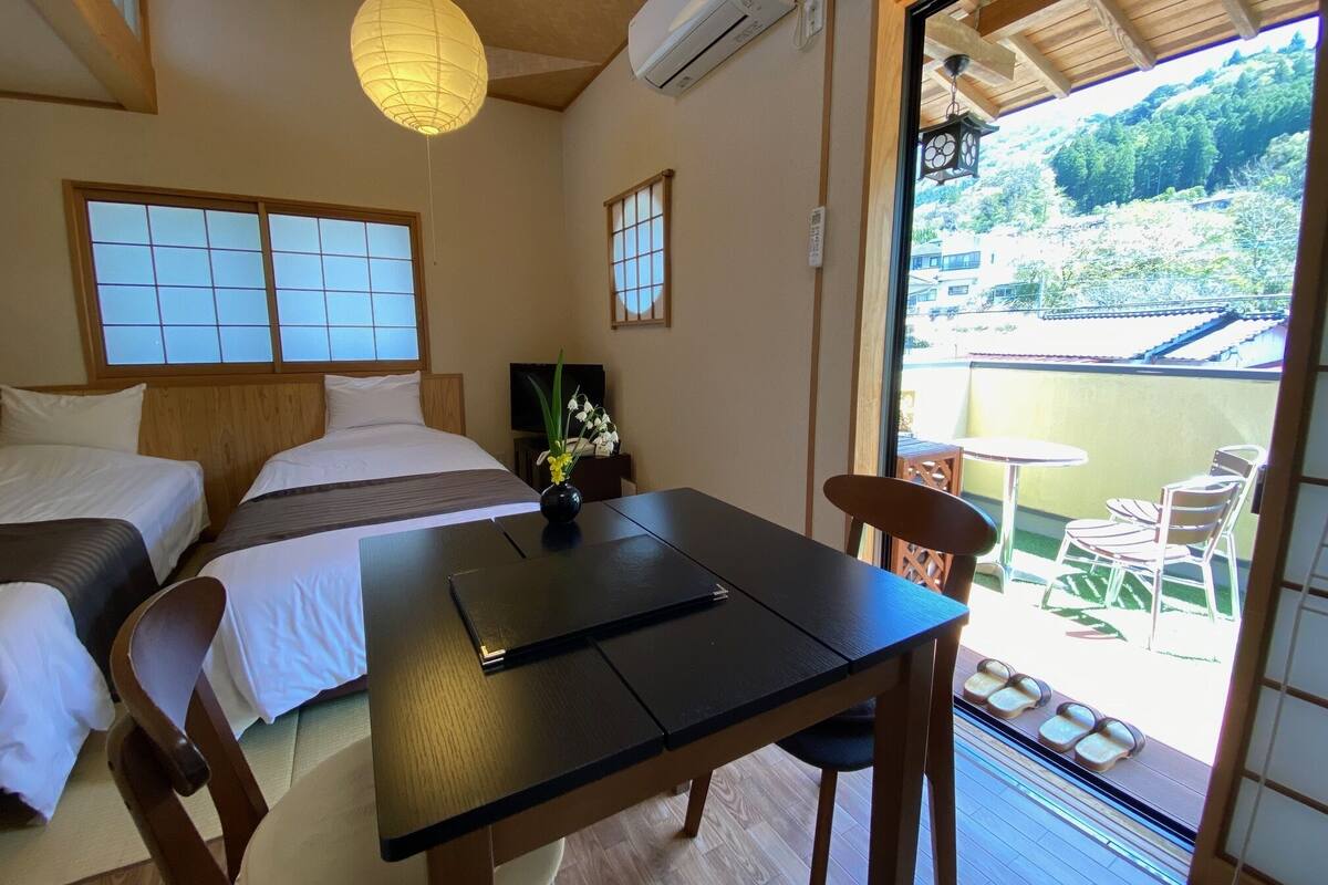 Quarto de hotel com duas camas, uma mesa de jantar, uma televisão e vista para um jardim através de uma porta de correr.