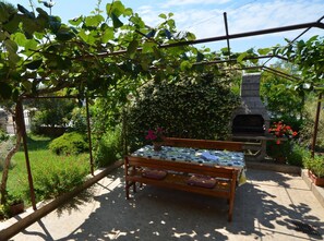 Um banco de madeira com almofadas sob uma pérgula coberta por plantas trepadeiras, um forno de pedra ao ar livre e um jardim com diversas plantas e flores.