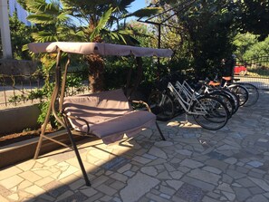 Um pátio com um banco estofado e um toldo, rodeado por plantas em vasos e uma fileira de bicicletas.