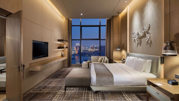 Quarto de hotel moderno com uma cama grande, televisão, secretária e vista para o horizonte urbano através de amplas janelas.