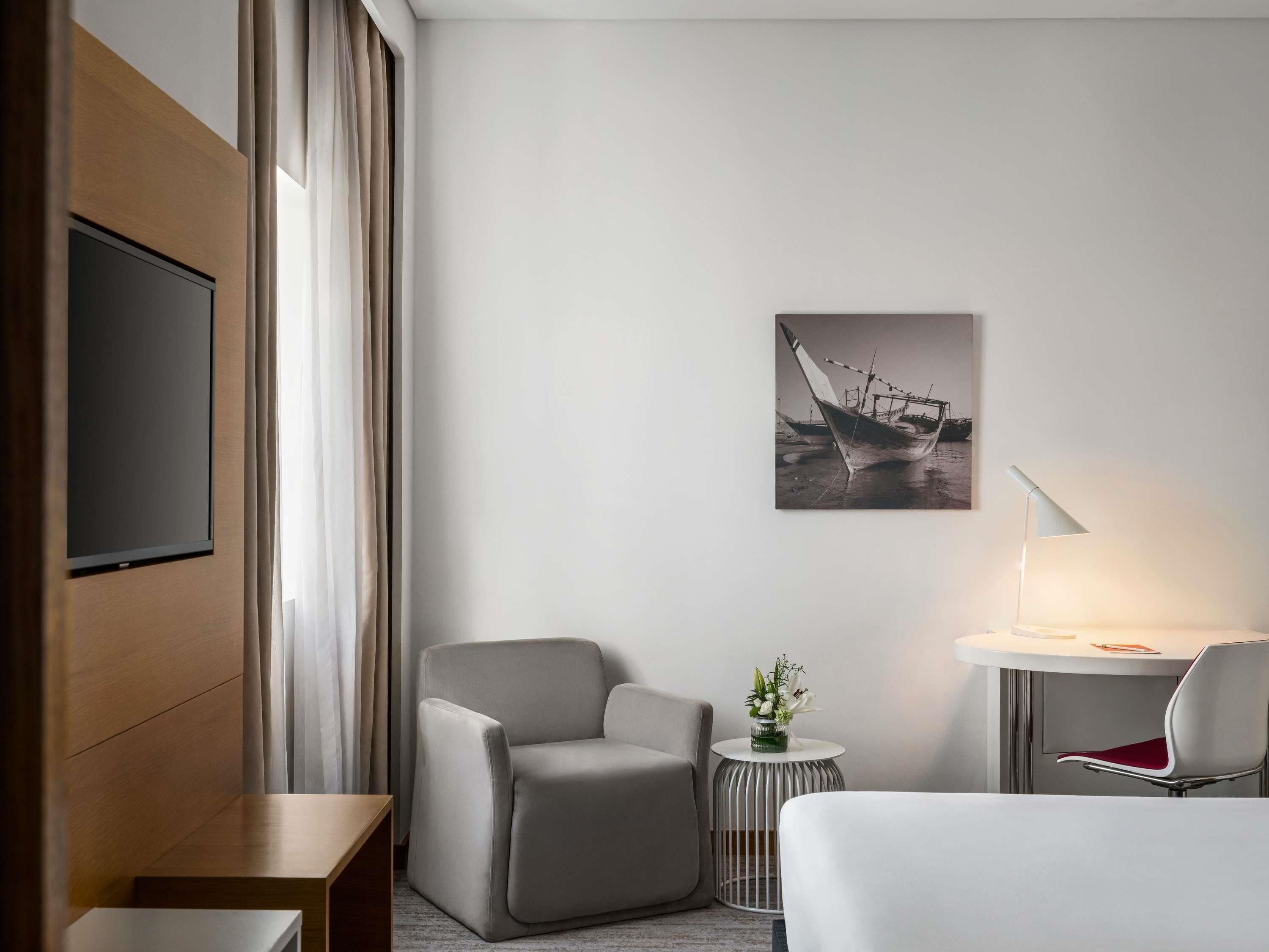 Quarto de hotel moderno com televisão de tela plana, uma poltrona confortável, uma mesinha redonda com um vaso de flores, uma escrivaninha com luminária e uma cama.