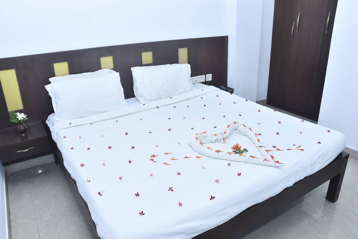 Uma cama bem arrumada com roupa de cama branca e padrão floral, uma mesa de cabeceira de madeira com um vaso, e um guarda-roupa de madeira escura.