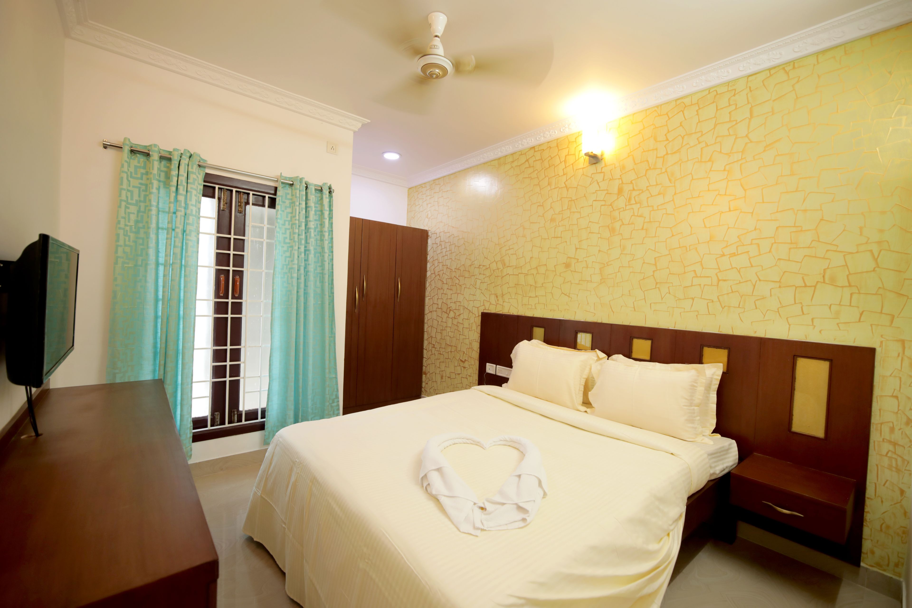 Quarto de hotel com cama, mesa de cabeceira de madeira, escrivaninha, televisão e janela com cortinas.