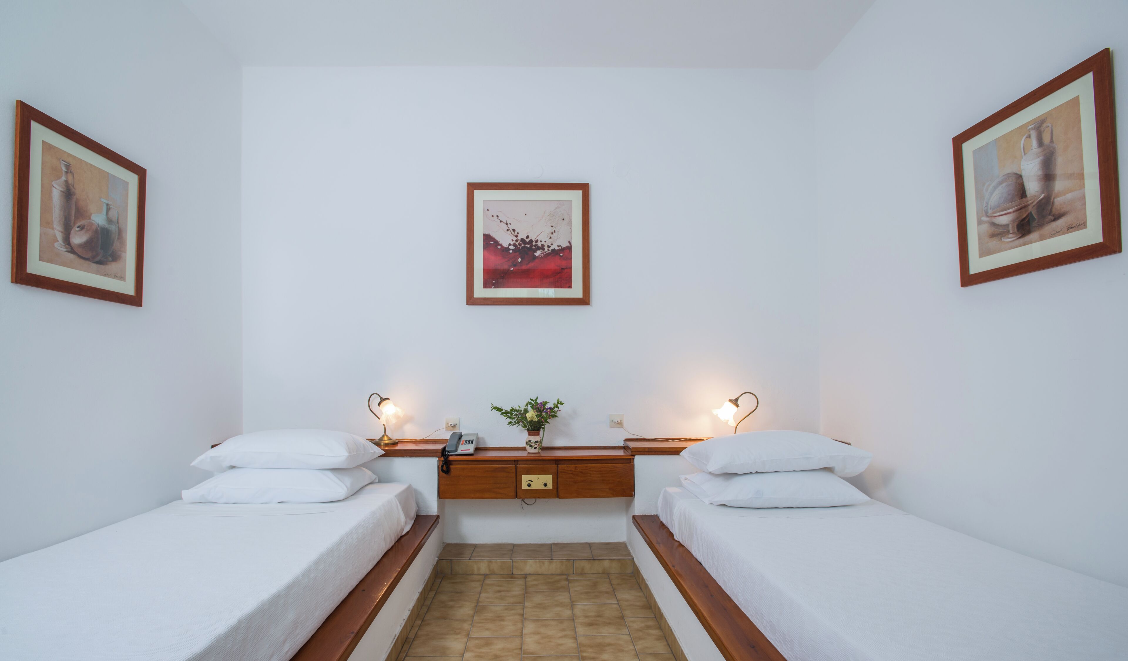Quarto com duas camas de solteiro, mesa de cabeceira de madeira com telefone, vaso com flores e quadros nas paredes.