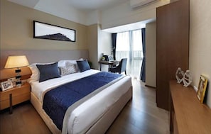 Quarto de hotel com cama, mesa de cabeceira, luminária e escrivaninha com cadeira.