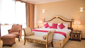 Quarto de hotel com uma cama grande, duas mesas de cabeceira com luminárias, uma cadeira e um banquinho.