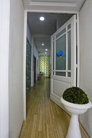Um corredor com piso de madeira, uma porta branca com painéis de vidro jateado e um pedestal branco com uma planta verde.