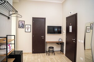 Um quarto pequeno e bem arrumado, com uma escrivaninha, duas portas, uma TV e um espelho.