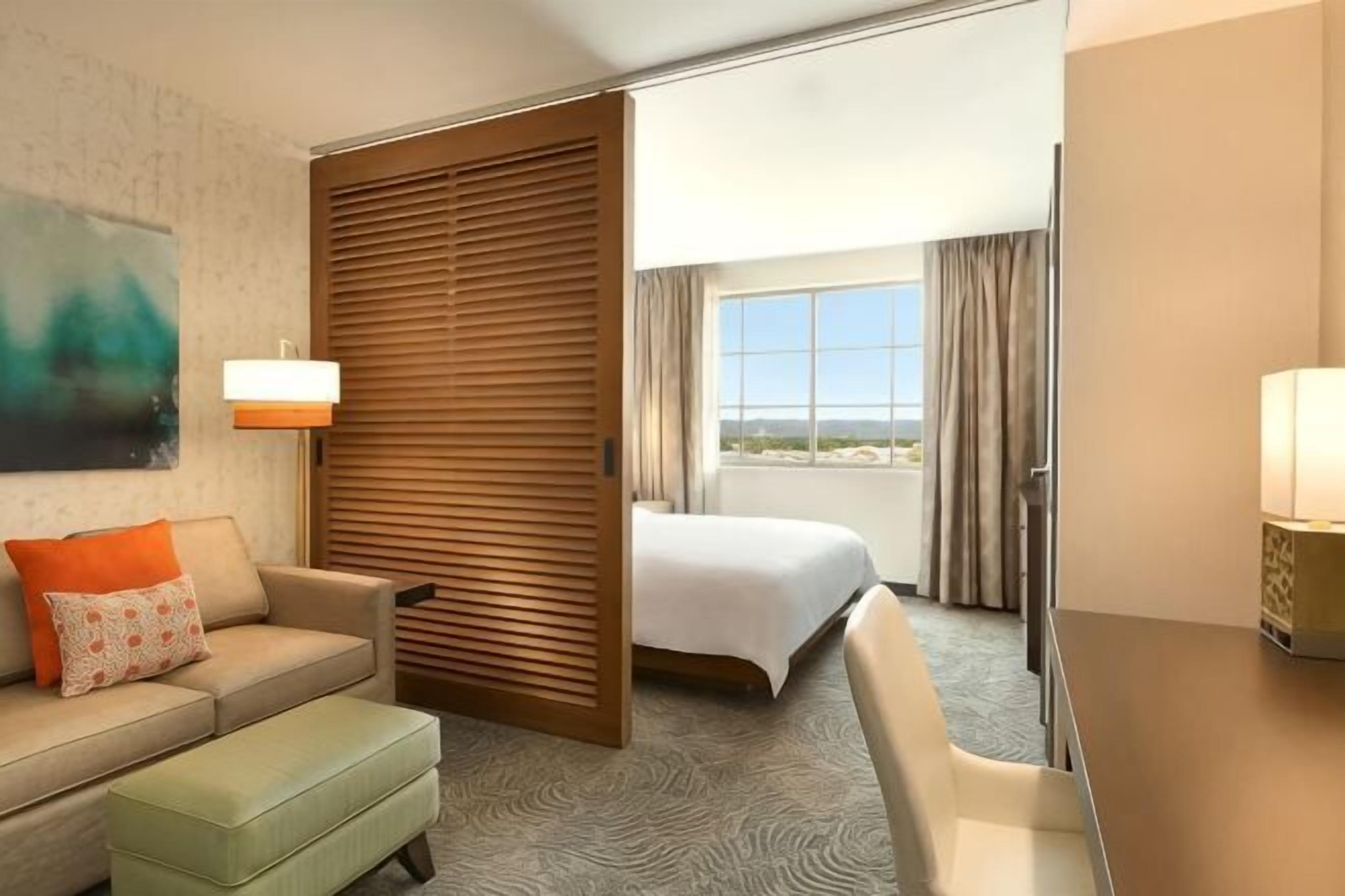 Quarto de hotel com cama, sofá, escrivaninha, luminária e janela com cortinas.
