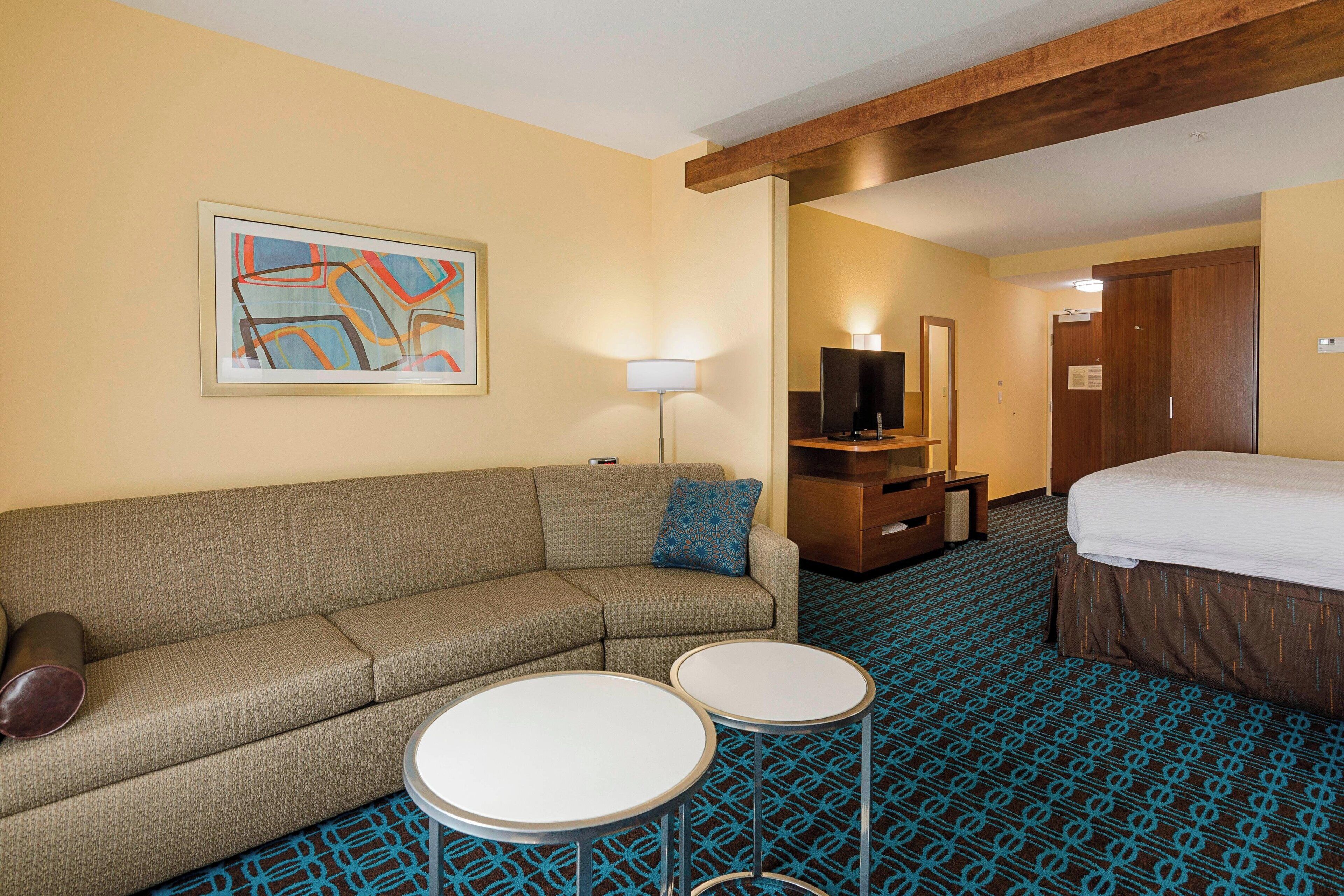 Quarto de hotel com sofá, duas mesinhas redondas, televisão, cama e tapete estampado.