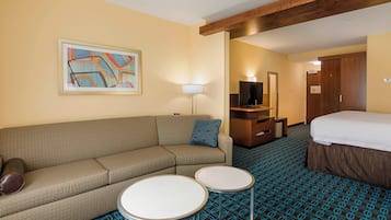 Quarto de hotel com sofá, duas mesinhas redondas, televisão, cama e tapete estampado.