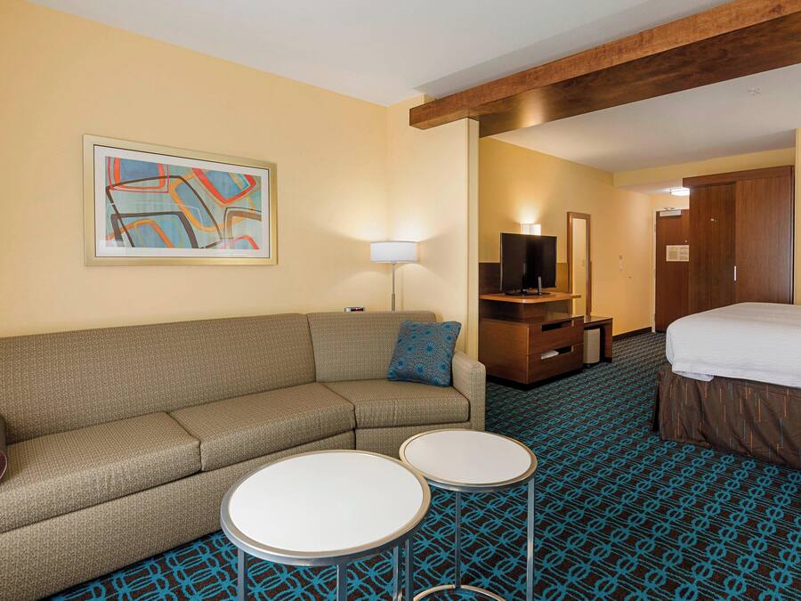 Quarto de hotel com sofá, duas mesinhas redondas, televisão, cama e tapete estampado.