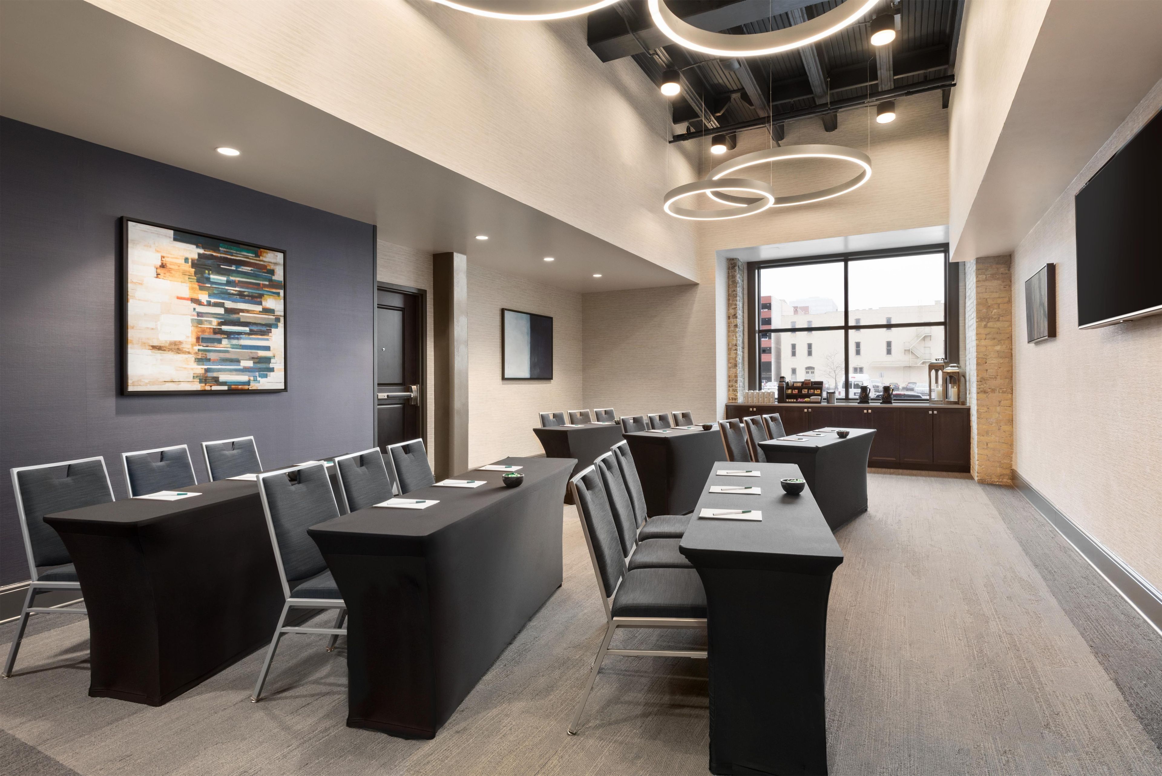 Sala de conferências com uma mesa comprida, cadeiras e uma janela grande com vista para edifícios.