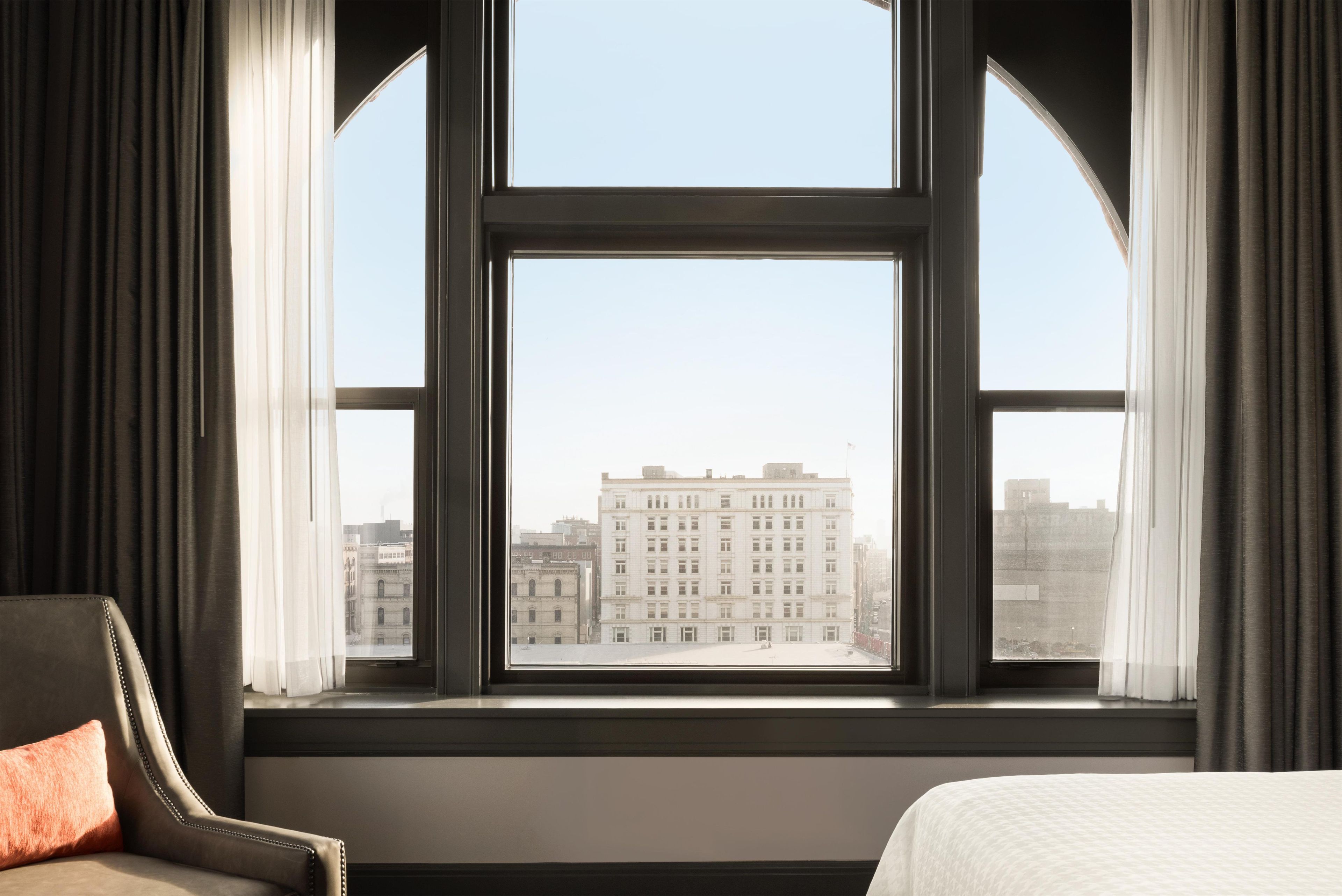 Quarto de hotel com uma grande janela em arco, uma cama, uma cadeira e vista para a cidade.