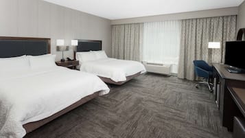Quarto de hotel com duas camas, uma escrivaninha com cadeira, televisão e janela com cortinas.