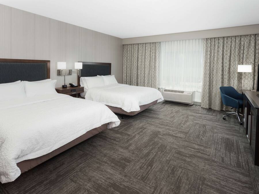Quarto de hotel com duas camas, uma escrivaninha com cadeira, televisão e janela com cortinas.