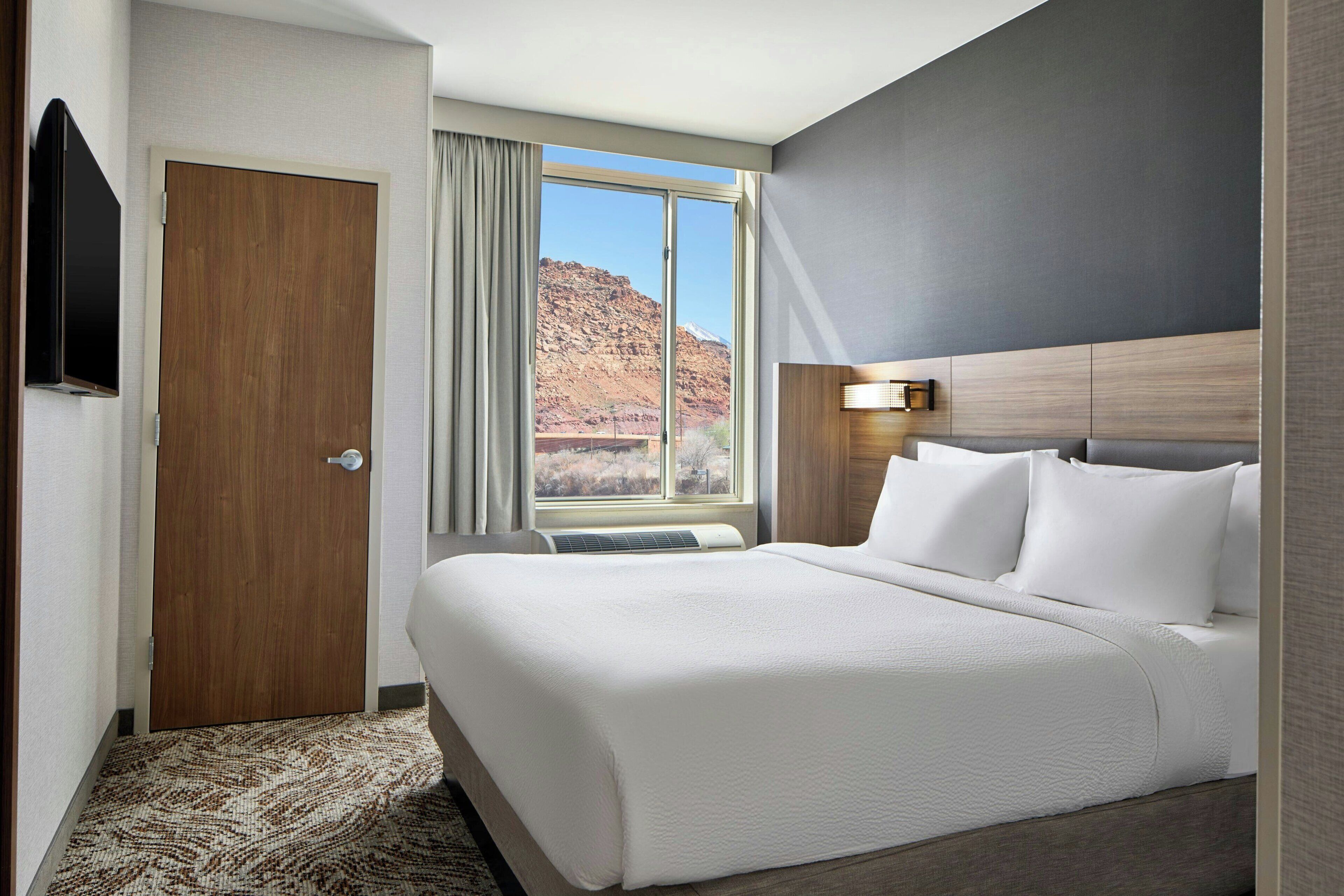 Quarto de hotel com uma cama grande, televisão na parede, janela com vista para uma montanha e uma porta.
